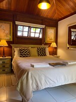 Cama bem arrumada com edredão branco e duas almofadas, mesa de cabeceira com abajur e janela com vista para o verde.