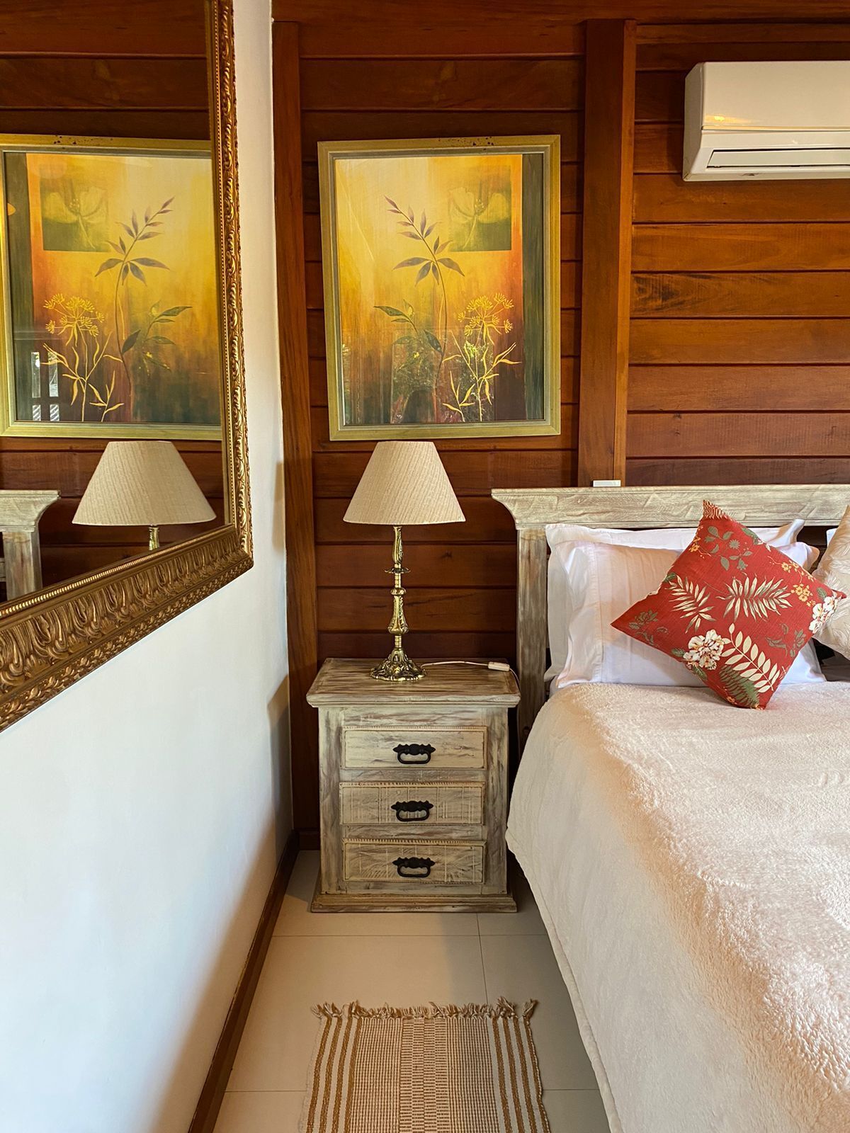 Quarto de hotel com parede de madeira, duas pinturas emolduradas, cama com roupa de cama branca e um travesseiro vermelho, mesa de cabeceira com abajur e uma pequena cômoda.