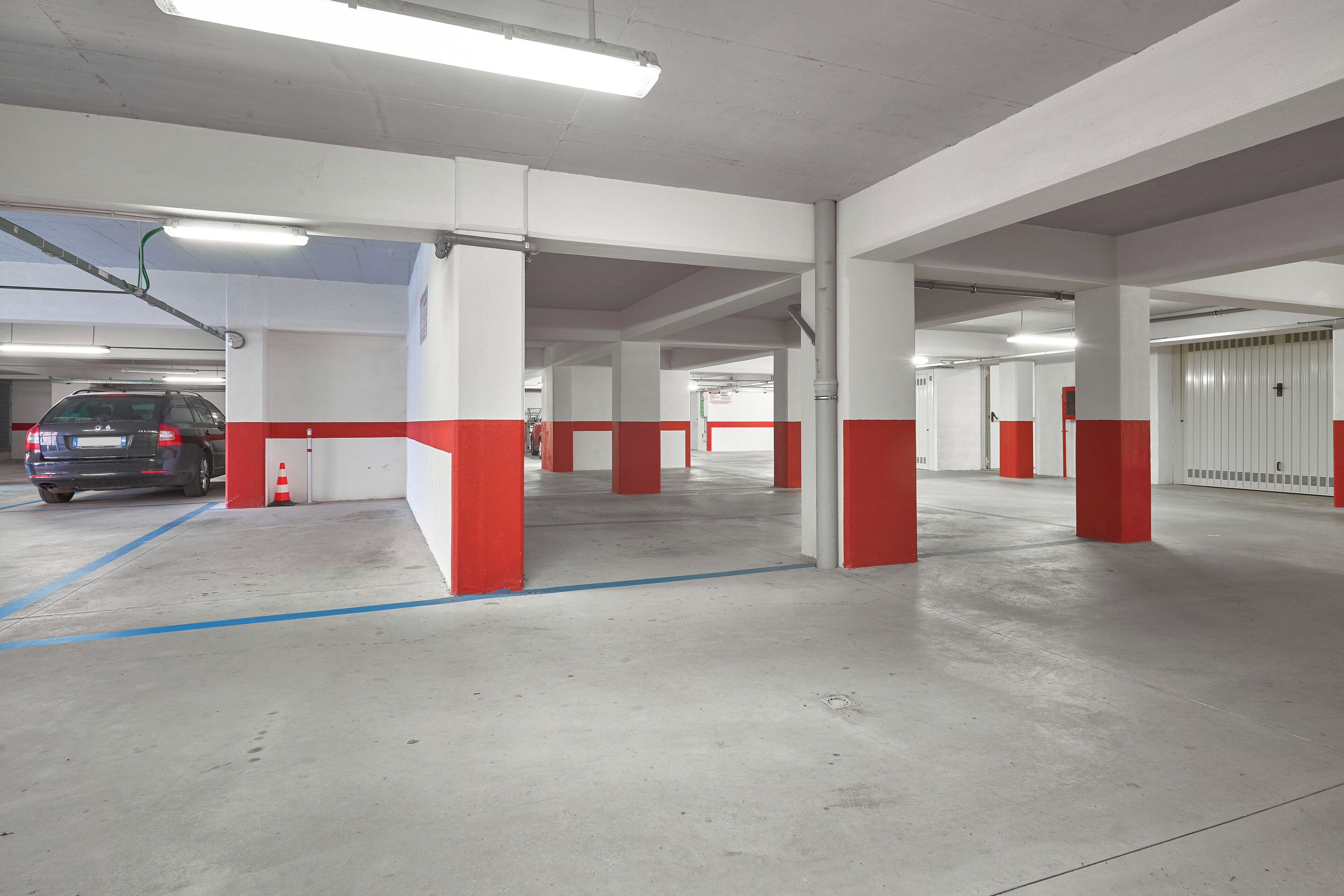 Um estacionamento multi-nível com pilares brancos e vermelhos, linhas azuis demarcando as vagas, e um carro estacionado ao fundo.