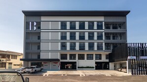 Um edifício moderno de vários andares com um logotipo 'B' em destaque, entrada coberta e área de estacionamento com vários carros.