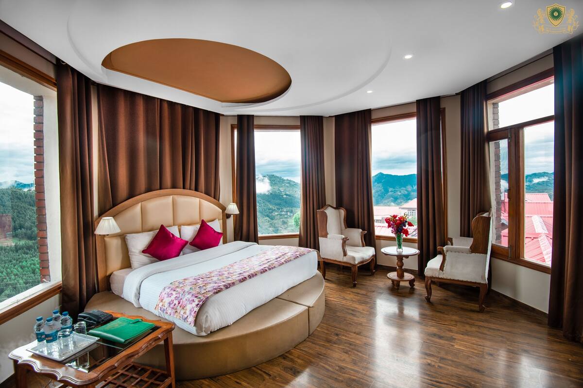 Quarto de hotel com uma cama grande, duas poltronas, uma mesinha e vista para as montanhas.