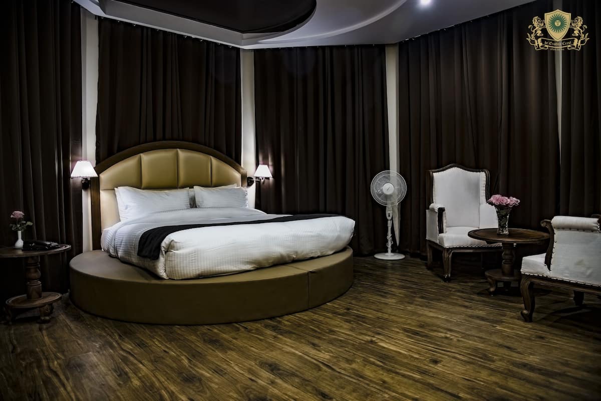 Quarto moderno com cama redonda, cortinas escuras, uma cadeira branca e uma mesinha com um vaso.