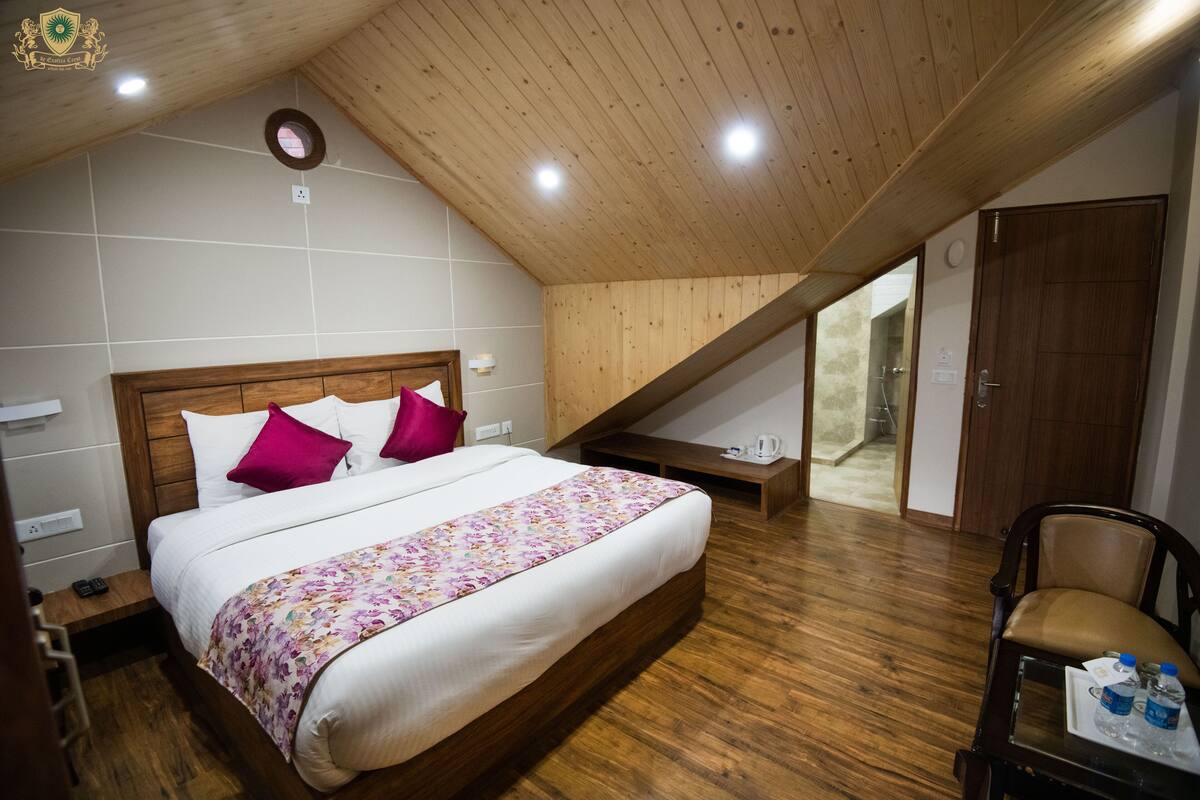 Quarto com teto de madeira, cama grande com roupa de cama branca e almofadas cor-de-rosa, um banquinho de madeira, uma cadeira e uma pequena mesa com uma garrafa e uma chávena.