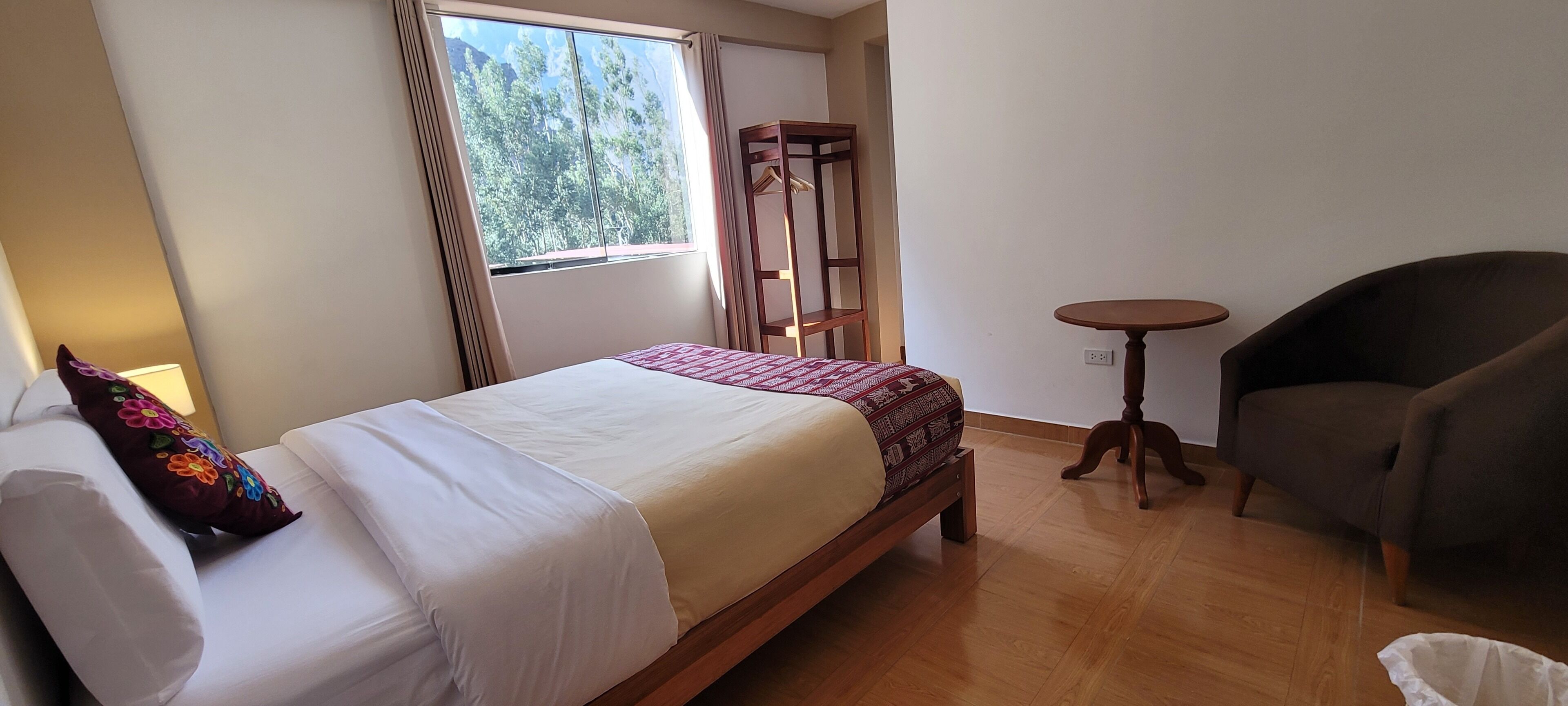 Quarto bem arrumado com cama, cadeira, mesa pequena e janela com vista para árvores.