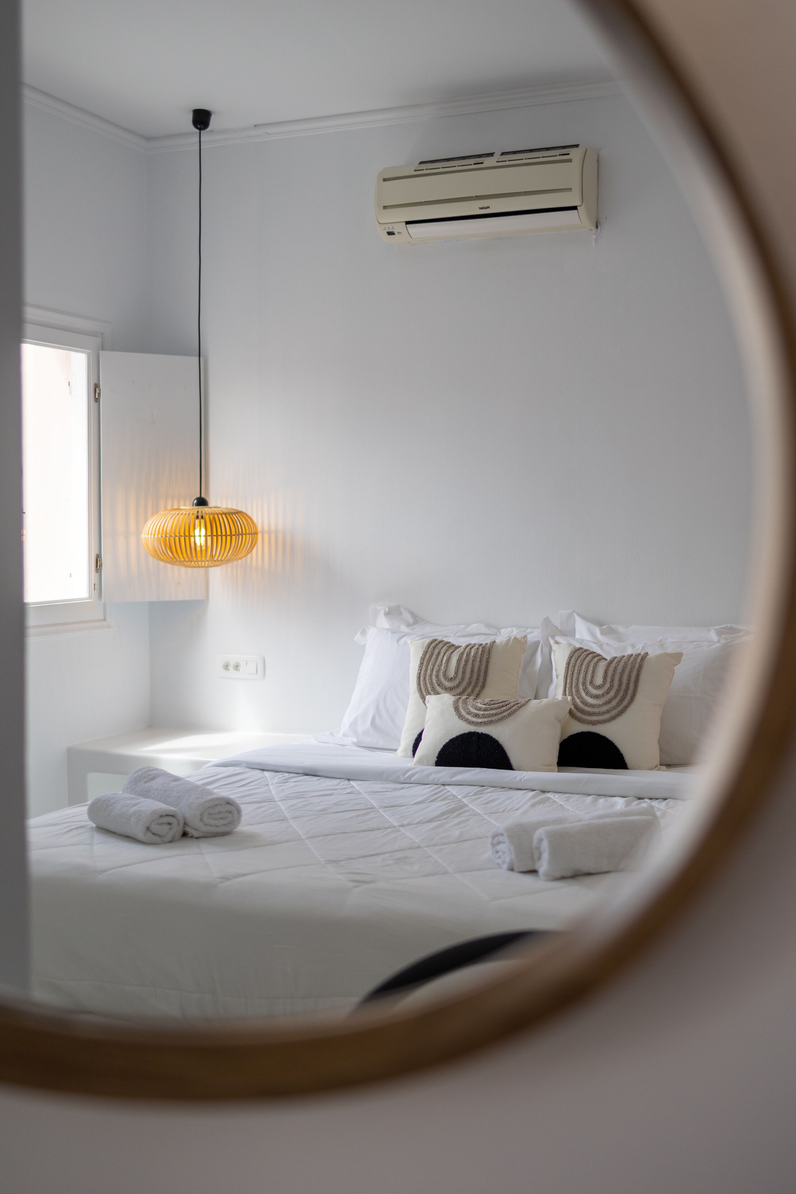 Quarto com cama, roupa de cama branca, luminária pendente e aparelho de ar condicionado.