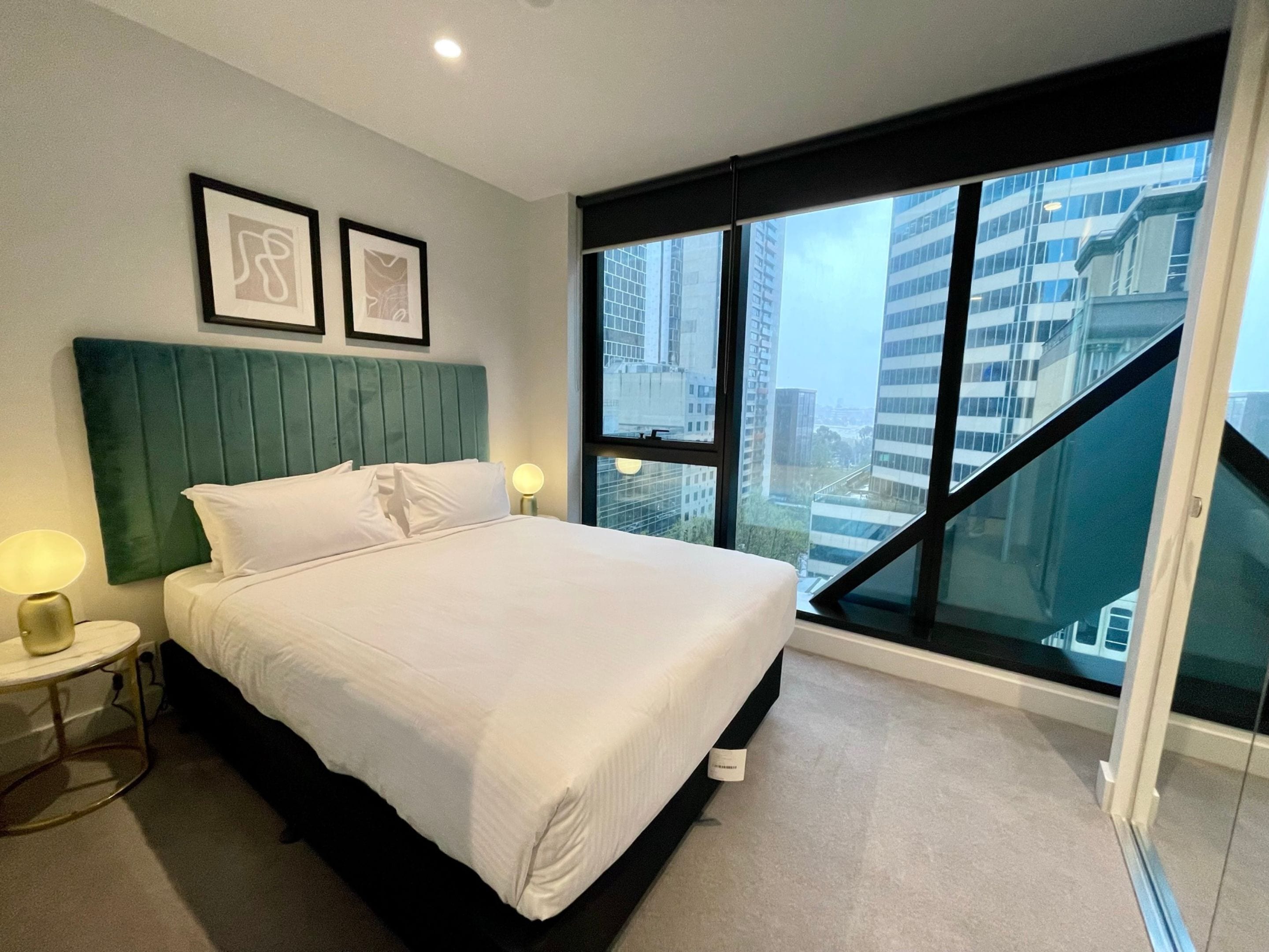 Quarto de hotel moderno com uma cama grande, duas obras de arte emolduradas na parede, um abajur e vista para a cidade através de uma janela ampla.