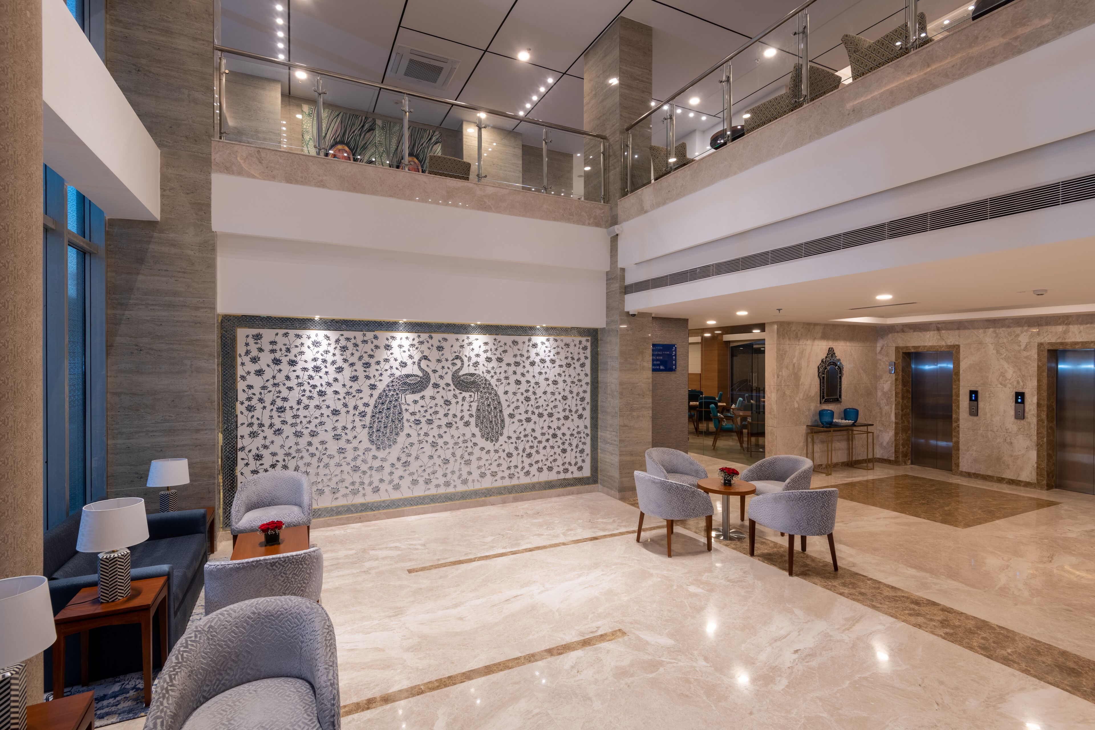 Um hall amplo com teto alto, uma parede decorativa com motivos de pavão e uma área de estar com cadeiras e mesas modernas.