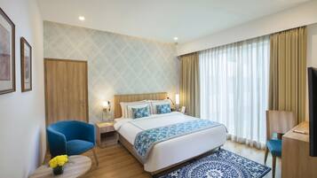 Quarto de hotel com cama, escrivaninha, cadeira, mesa pequena com um vaso e uma janela grande com cortinas.