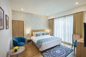 Quarto de hotel com cama, escrivaninha, cadeira, mesa pequena com um vaso e uma janela grande com cortinas.