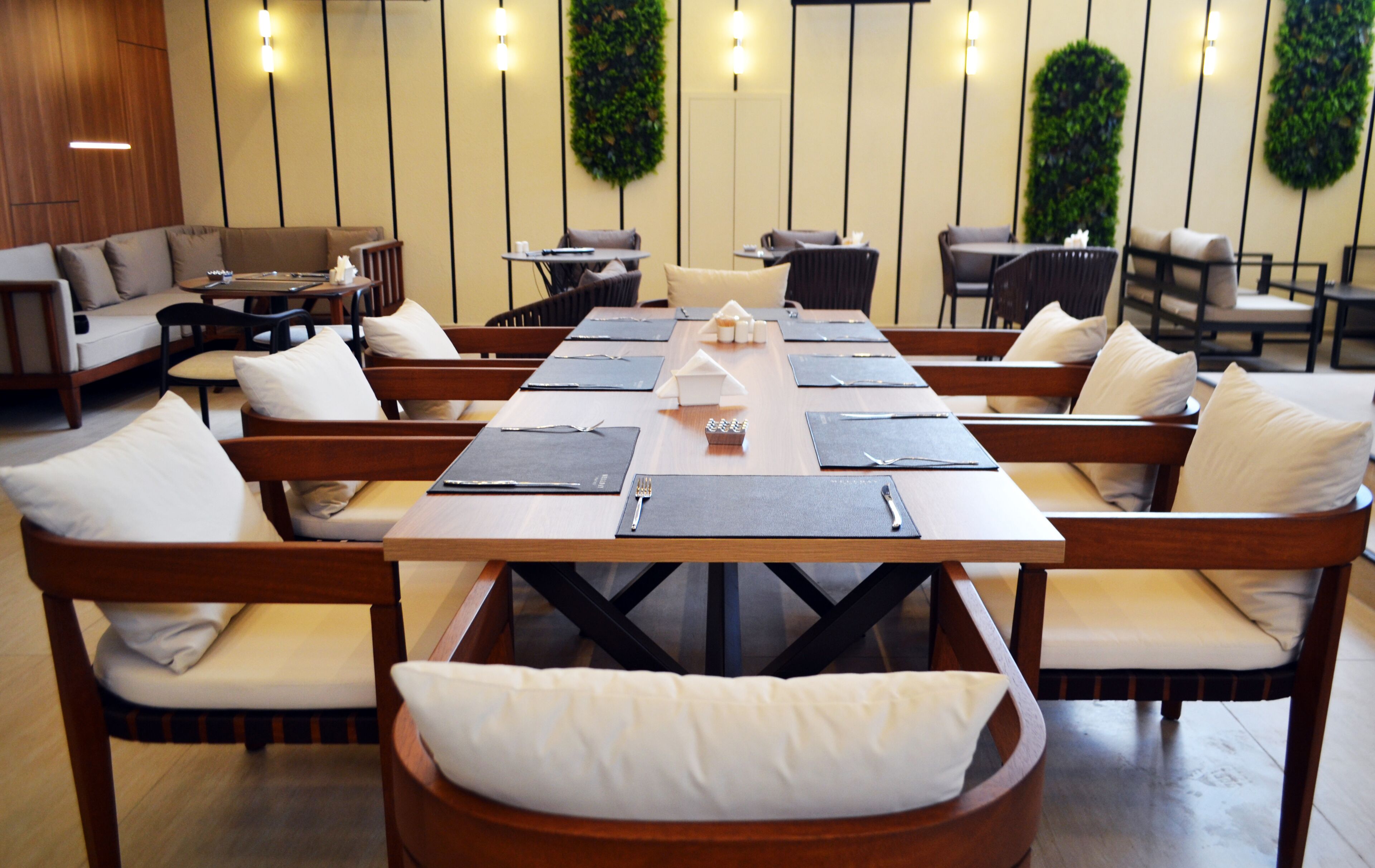 Uma sala de jantar com uma longa mesa de madeira posta para seis pessoas, rodeada por cadeiras de madeira com almofadas brancas.