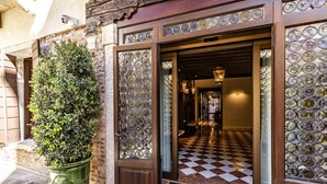 Entrada de hotel com chão xadrez, tapete de boas-vindas verde com um logotipo e uma planta em vaso.