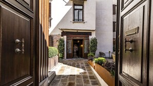 Um caminho de pedra estreito que leva à entrada de um edifício com portas de madeira maciça e um pequeno jardim com plantas em vasos.