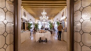 Um hall amplo com uma mesa central posta para refeição, cercada por plantas em vasos e um candelabro.