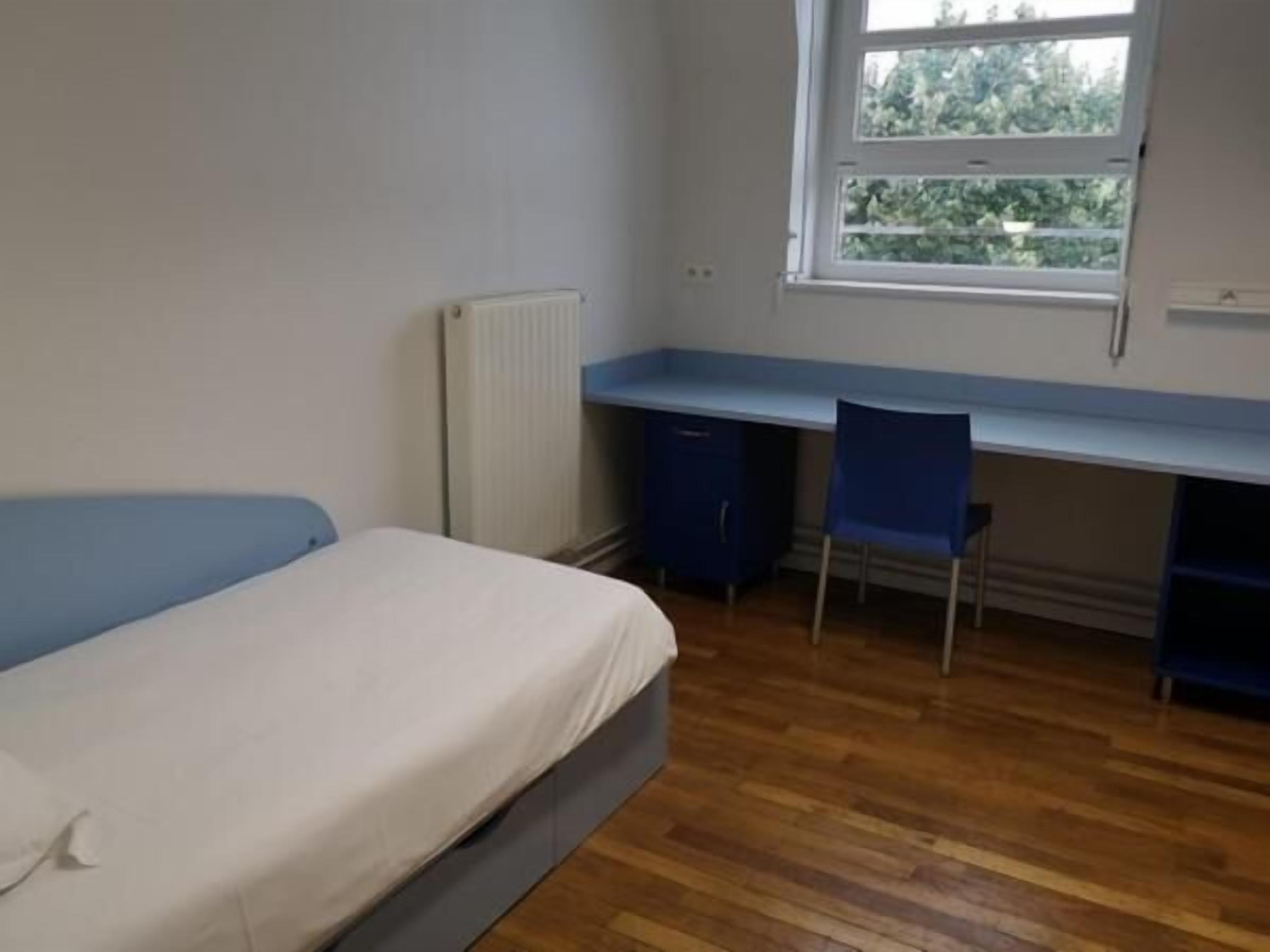 Um quarto com uma cama, uma escrivaninha, uma cadeira e um radiador.