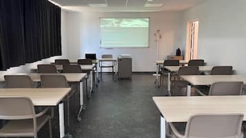 Sala de conferências com tela de projeção exibindo uma apresentação, várias mesas e cadeiras, e uma cortina preta em um dos lados.