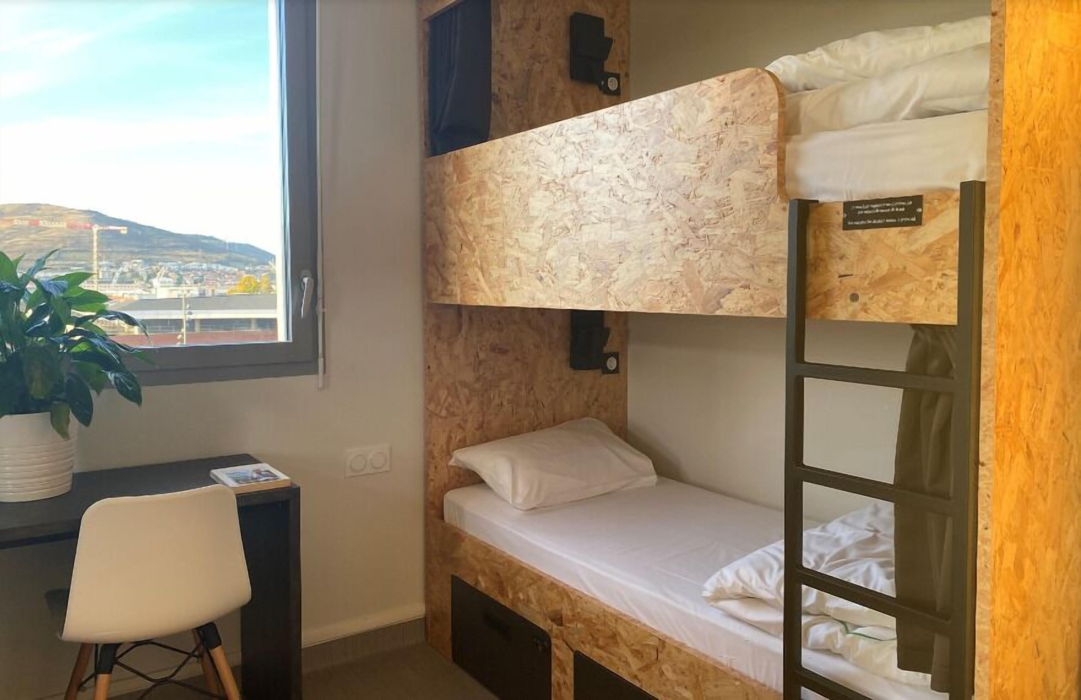Quarto com beliche e vista para a cidade, uma escrivaninha com cadeira e um vaso de planta.