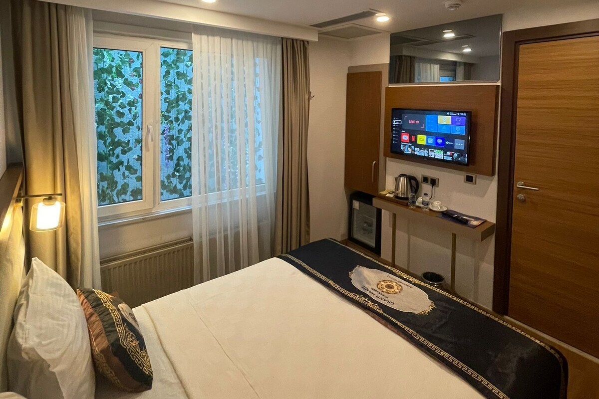 Quarto de hotel com cama, televisão, escrivaninha com cafeteira e janela com cortinas.