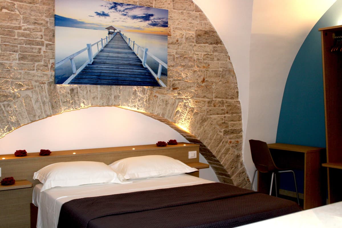 Quarto com parede de pedra, cabeceira de madeira, cama com roupa de cama branca e um quadro de um cais.