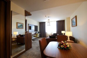 Quarto de hotel com mesa de jantar de madeira, duas poltronas, um sofá, escrivaninha com computador, televisão e uma janela grande com cortinas.