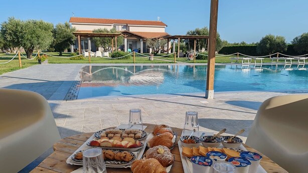 Mesa de refeições à beira da piscina, com diversos doces, frutas e bebidas, com vista para uma piscina e um edifício com telhado em azulejos.