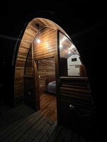 Interior acolhedor de uma cabana de madeira, com uma cama, uma mesa pequena e uma porta que dá para outro quarto.
