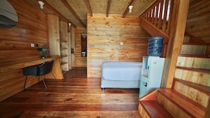 Interior de uma cabana de madeira com uma cama, uma escrivaninha, uma cadeira e um bebedouro.