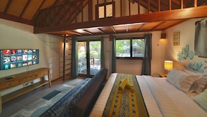 Quarto de hotel com uma cama grande, um banco de madeira, uma TV fixada na parede e uma varanda com vista para o exterior.