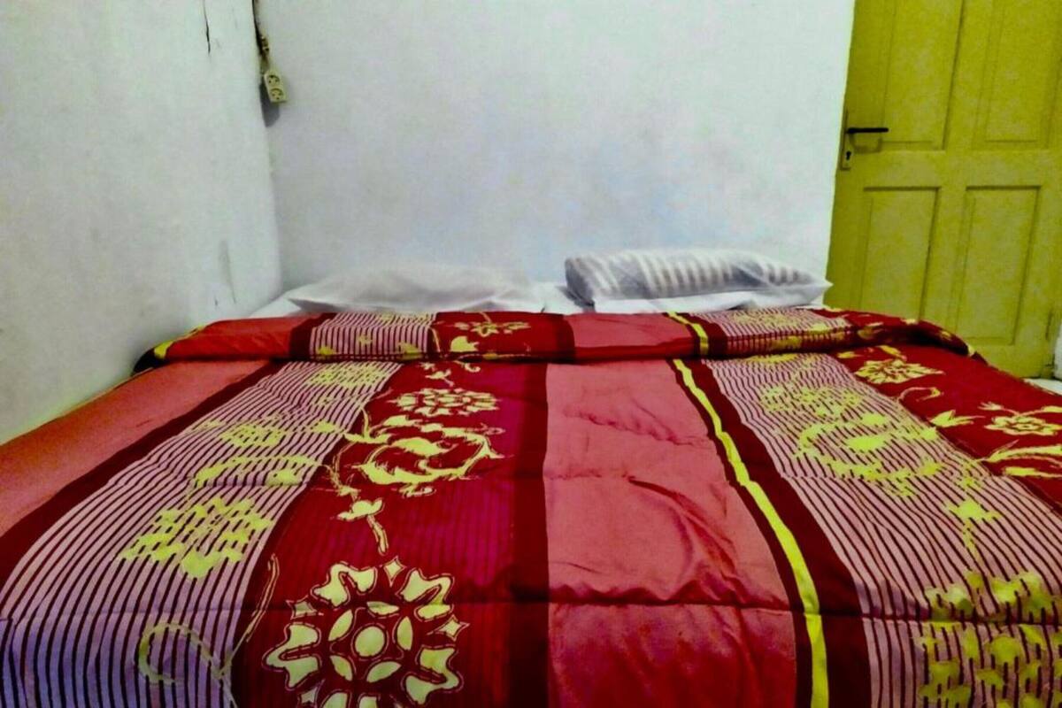 Uma cama com um cobertor de padrão vermelho e branco, um travesseiro branco e uma porta amarela ao fundo.