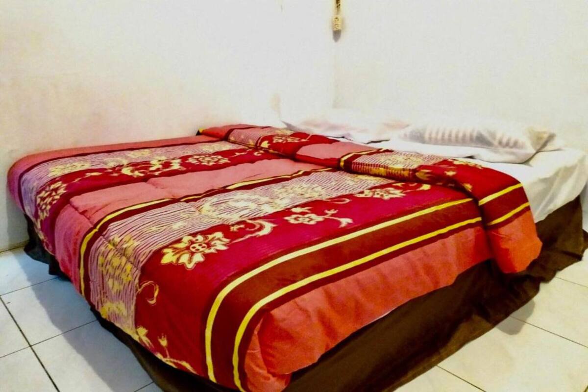 Uma cama de casal com roupa de cama vermelha e dourada, almofadas brancas e parede branca.