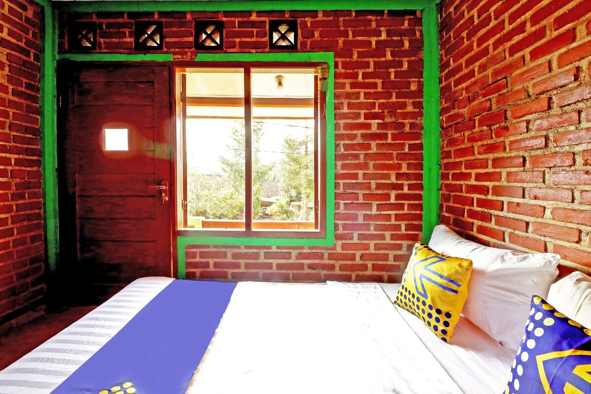 Quarto com parede de tijolos, porta de madeira, janela com moldura verde e cama com roupa de cama branca e azul.
