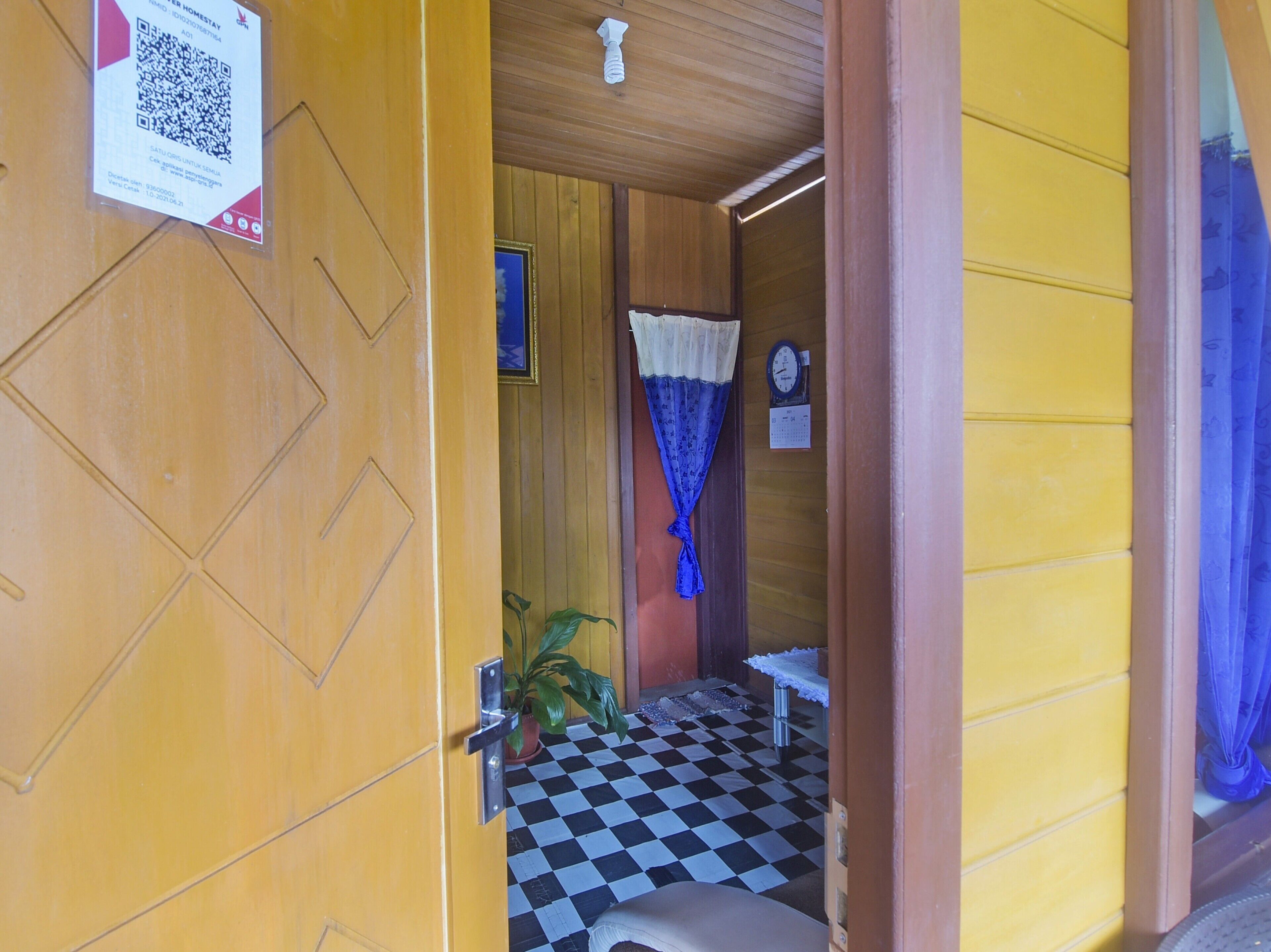 Quarto com piso xadrez, porta de madeira, uma planta, um relógio e um calendário na parede.