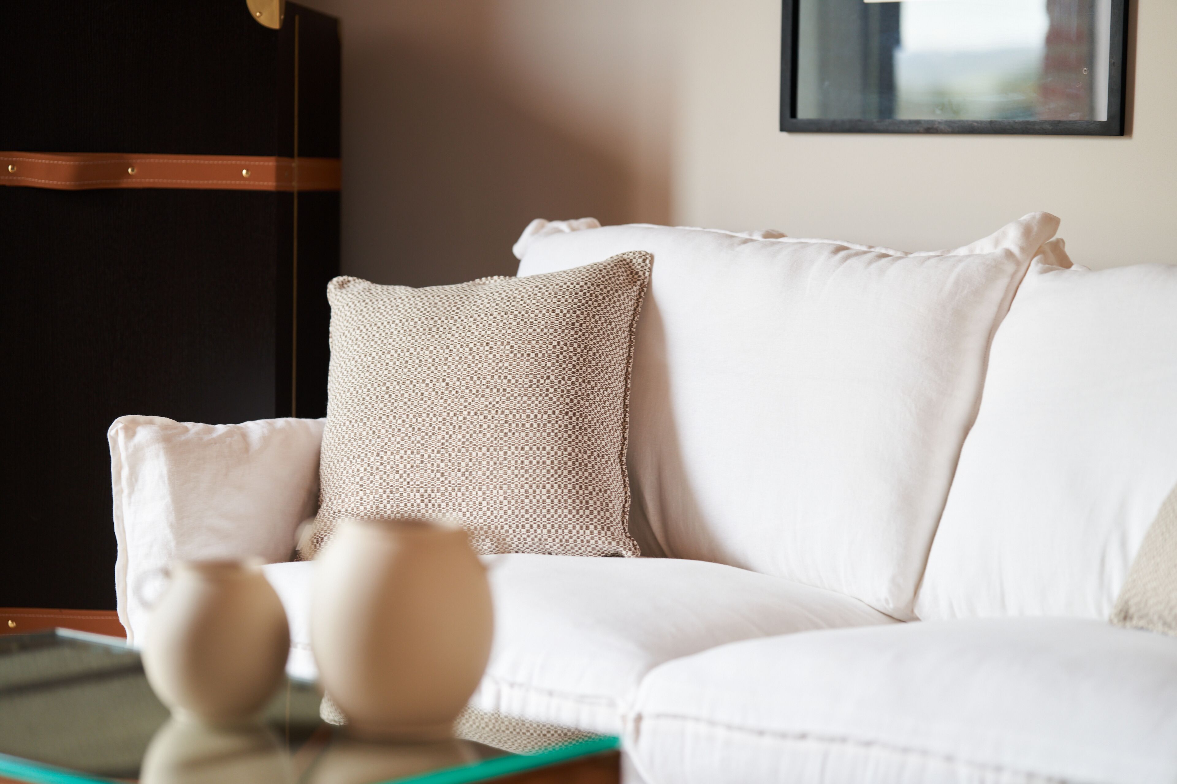 Um sofá branco com uma almofada texturizada e um travesseiro, posicionado em frente a uma parede com um quadro emoldurado.
