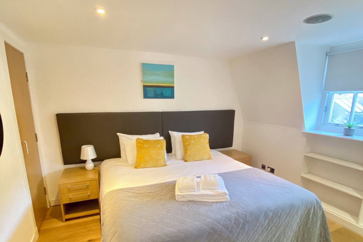 Quarto com cama, mesa de cabeceira, abajur e um quadro na parede.