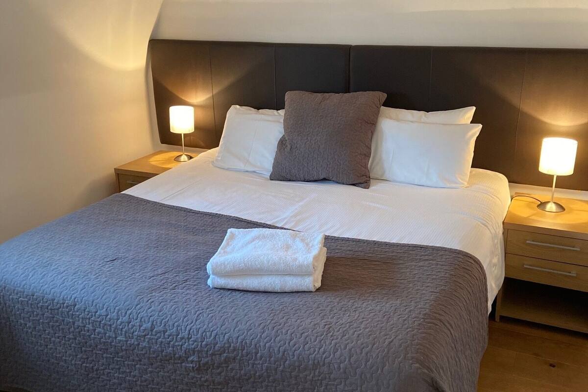 Cama bem arrumada com manta cinza, duas almofadas e uma toalha dobrada. Há duas mesas de cabeceira com abajures de cada lado da cama.