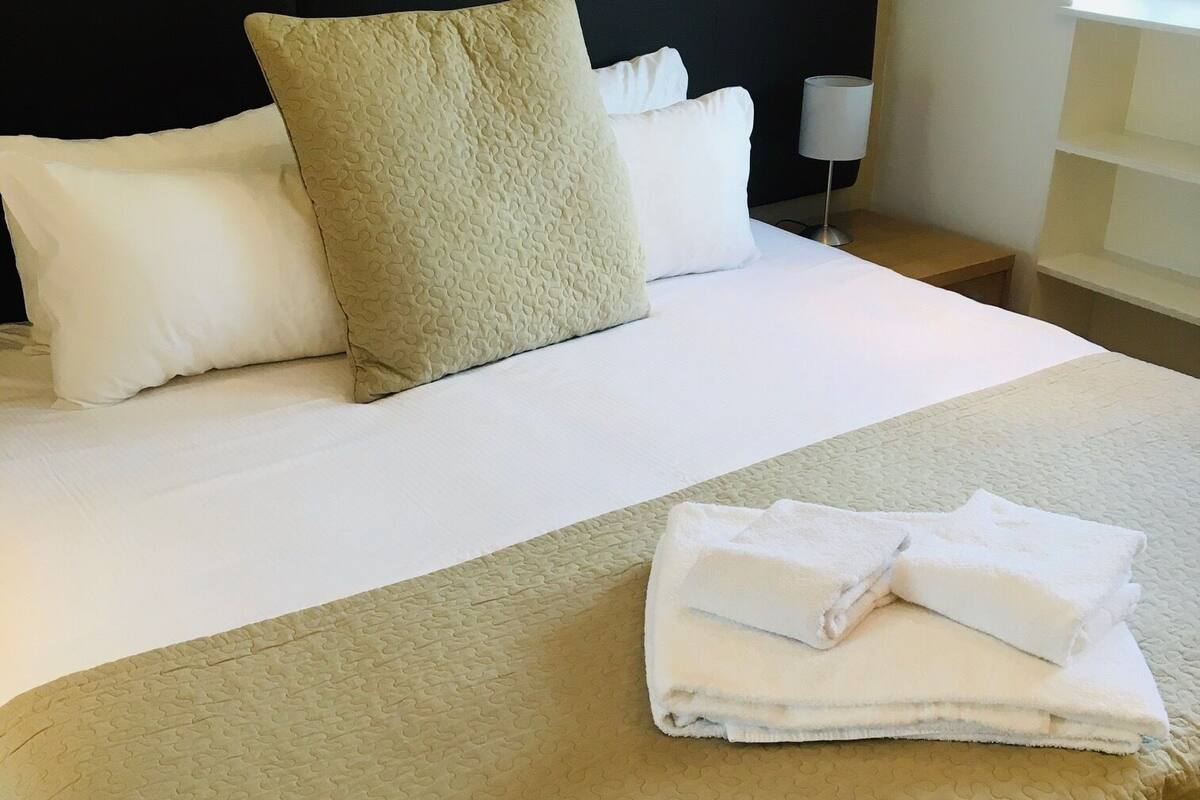 Cama bem arrumada com colcha bege, almofadas brancas e uma toalha branca dobrada.