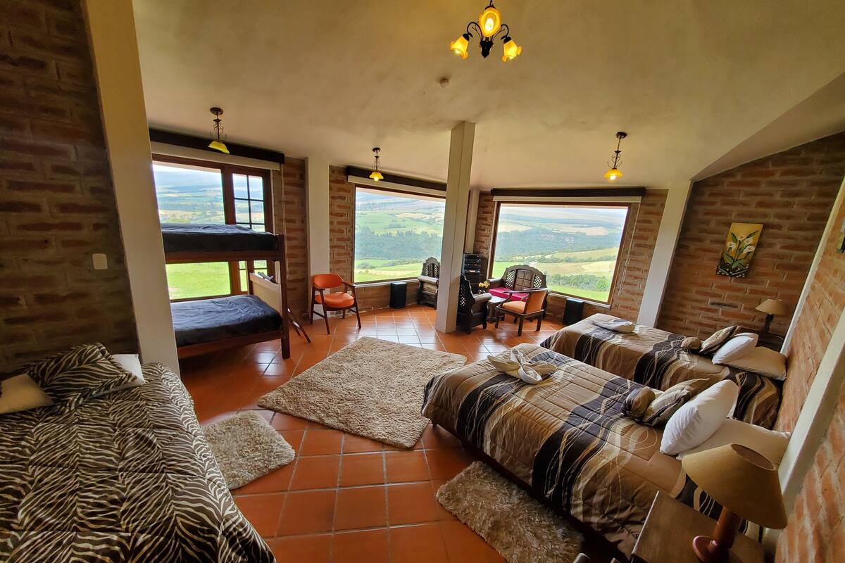 Quarto espaçoso com uma cama grande, uma cama menor e um sofá. Há uma mesa e uma cadeira no centro, e vista para o campo através de janelas amplas.
