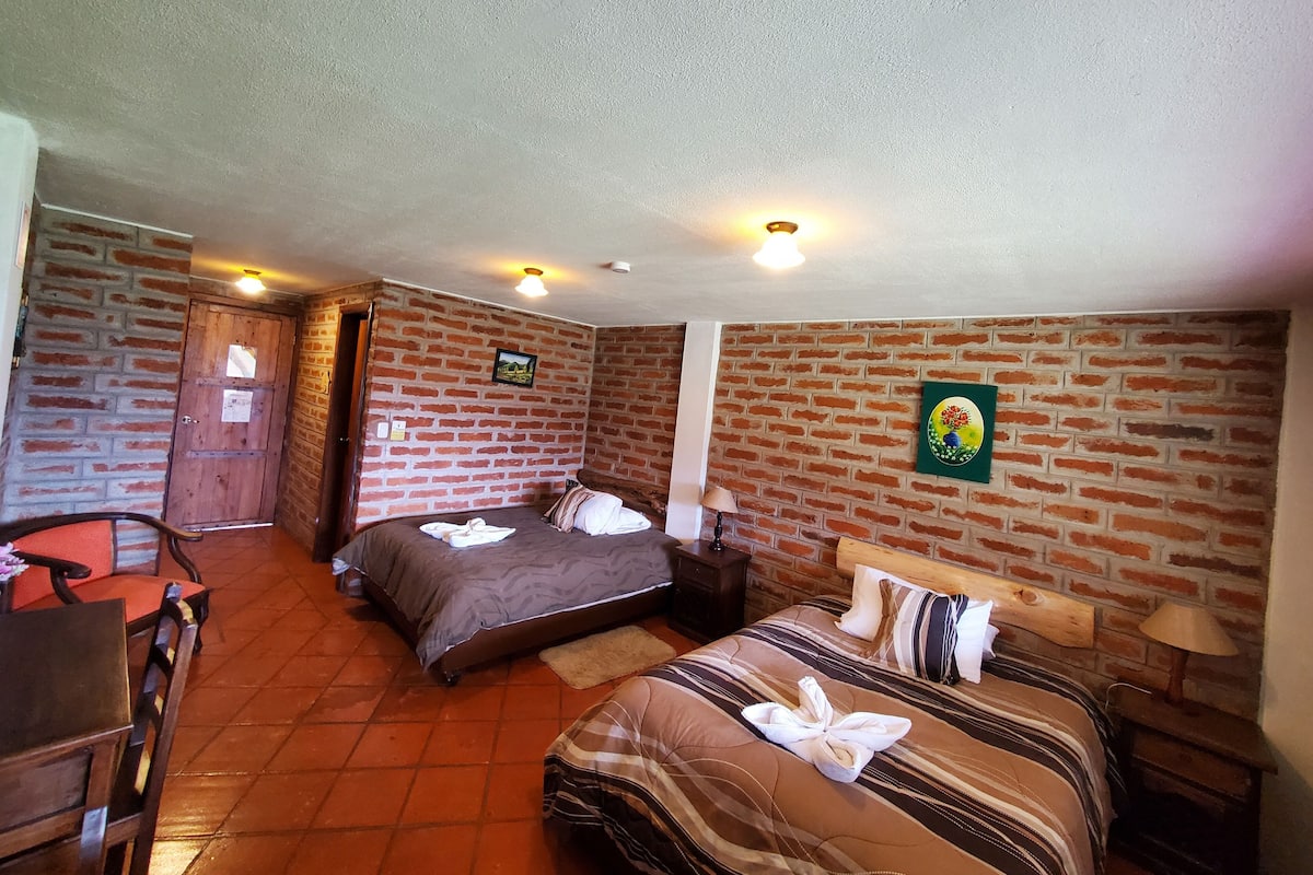 Um quarto com duas camas, uma cadeira de madeira, uma mesinha e uma parede de tijolos.