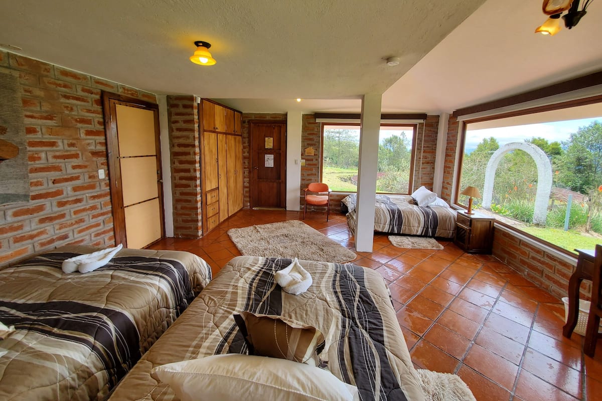 Quarto espaçoso com uma cama grande, uma cama menor e uma cadeira. Há uma janela com vista para o verde lá fora.