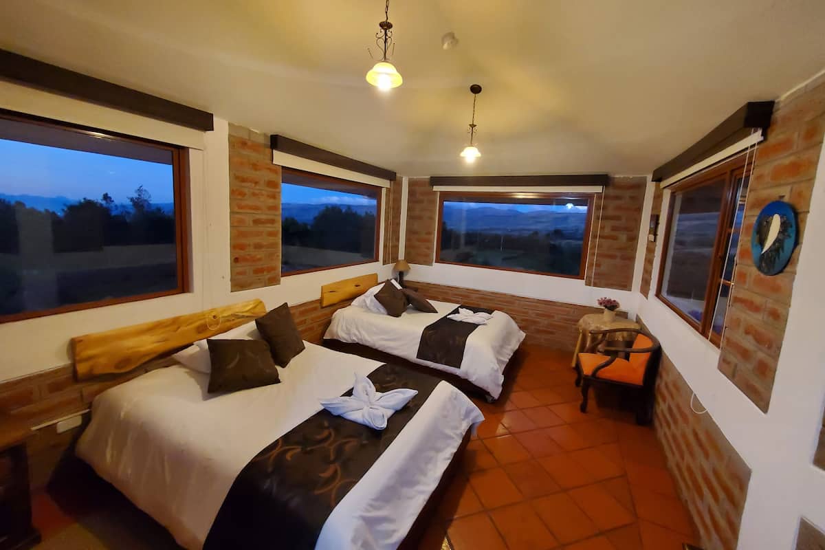 Quarto com duas camas, uma mesinha e janelas amplas com vista para a paisagem.