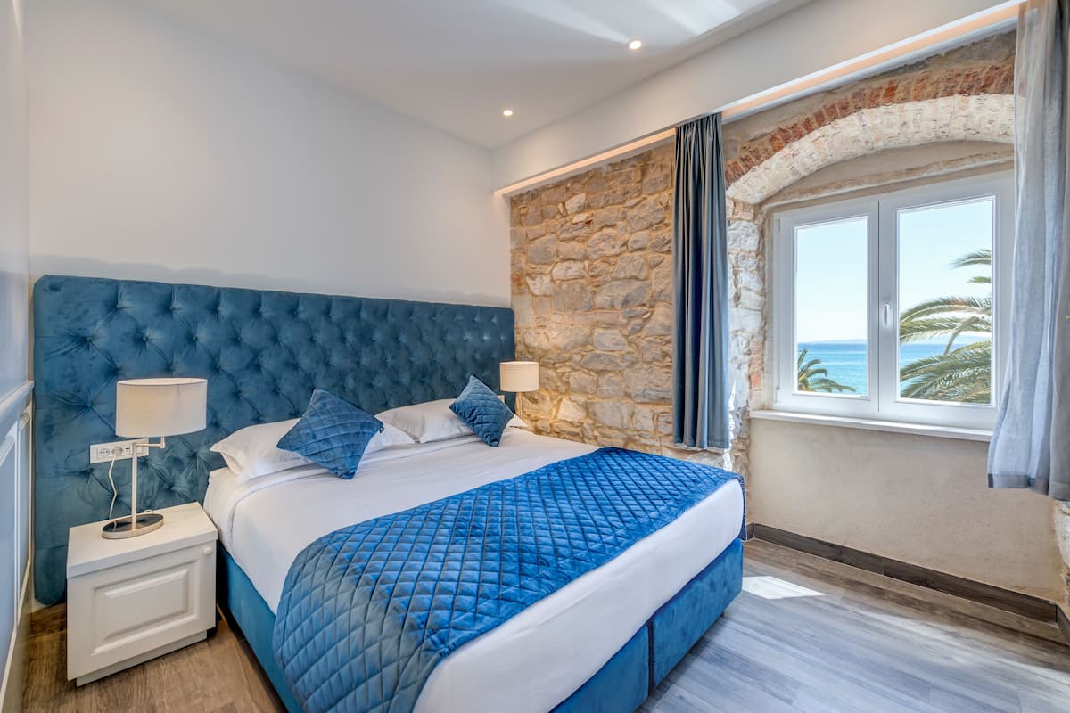 Quarto com parede de pedra, uma cama grande com colcha azul e vista para o mar pela janela.