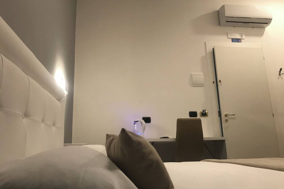 Quarto de hotel com cama, uma cadeira, escrivaninha e ar condicionado na parede.