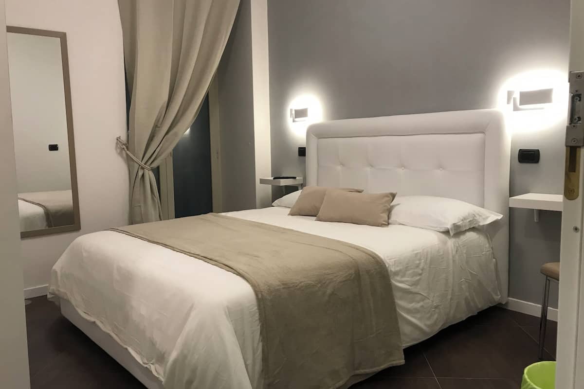 Cama bem arrumada com cabeceira branca, manta bege e duas almofadas.