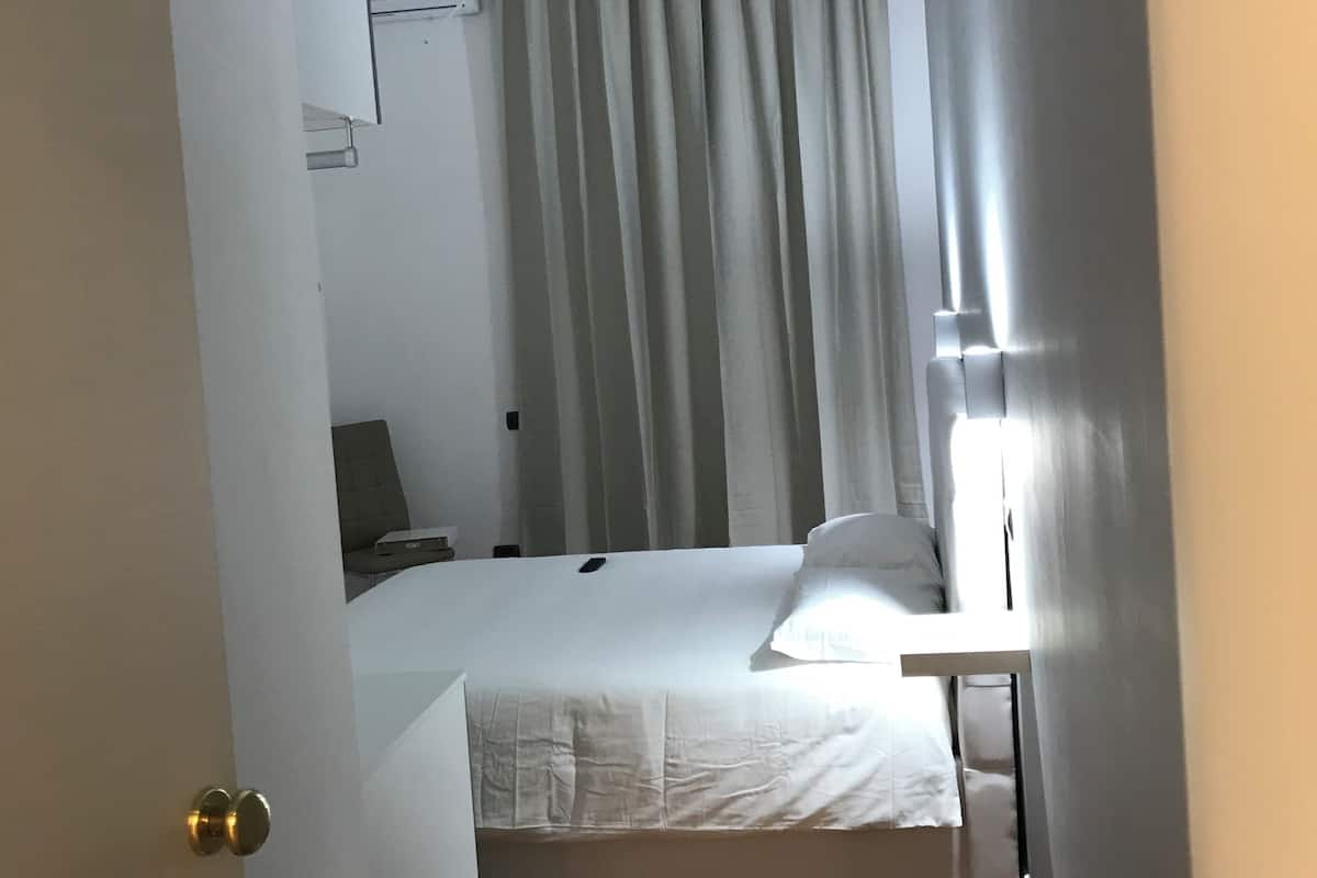 Quarto com cama, mesa de cabeceira, luminária e janela com cortinas.