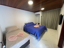 Quarto de hotel com duas camas, uma com cobertor rosa e outra com cobertor azul. Há um ventilador de teto, ar-condicionado fixado na parede e uma cortina na janela.
