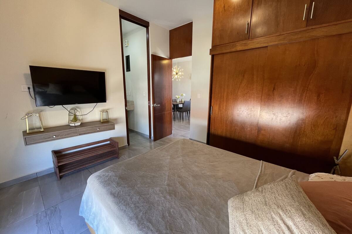 Quarto com cama grande, televisão na parede, guarda-roupa de madeira e uma pequena mesa com objetos decorativos.