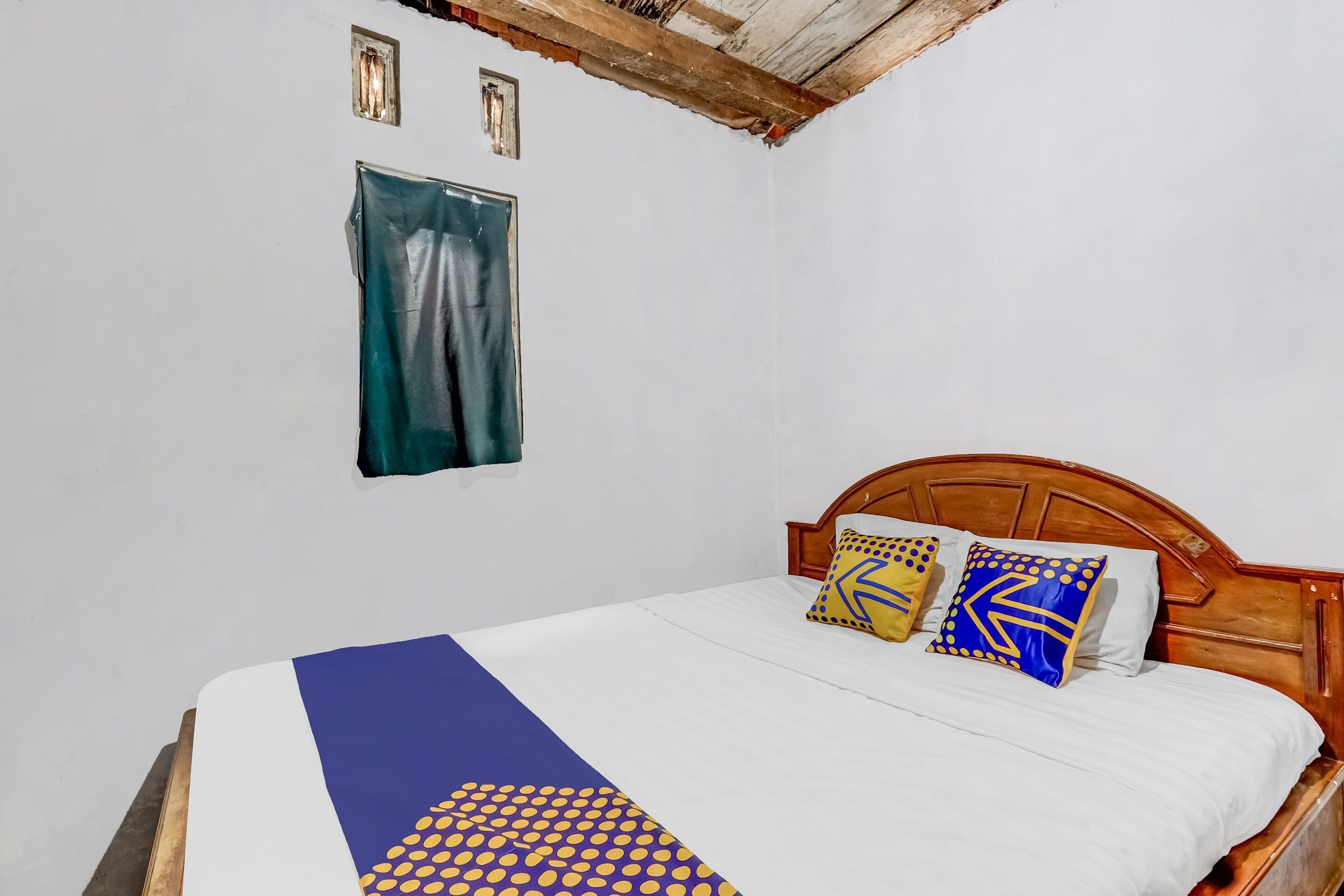 Quarto com cabeceira de madeira, cama com roupa de cama branca, colcha azul e amarela estampada e dois travesseiros decorativos.