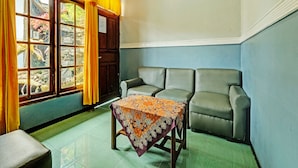 Um quarto com um sofá verde, uma mesa pequena com toalha de mesa estampada e uma janela com cortinas amarelas.