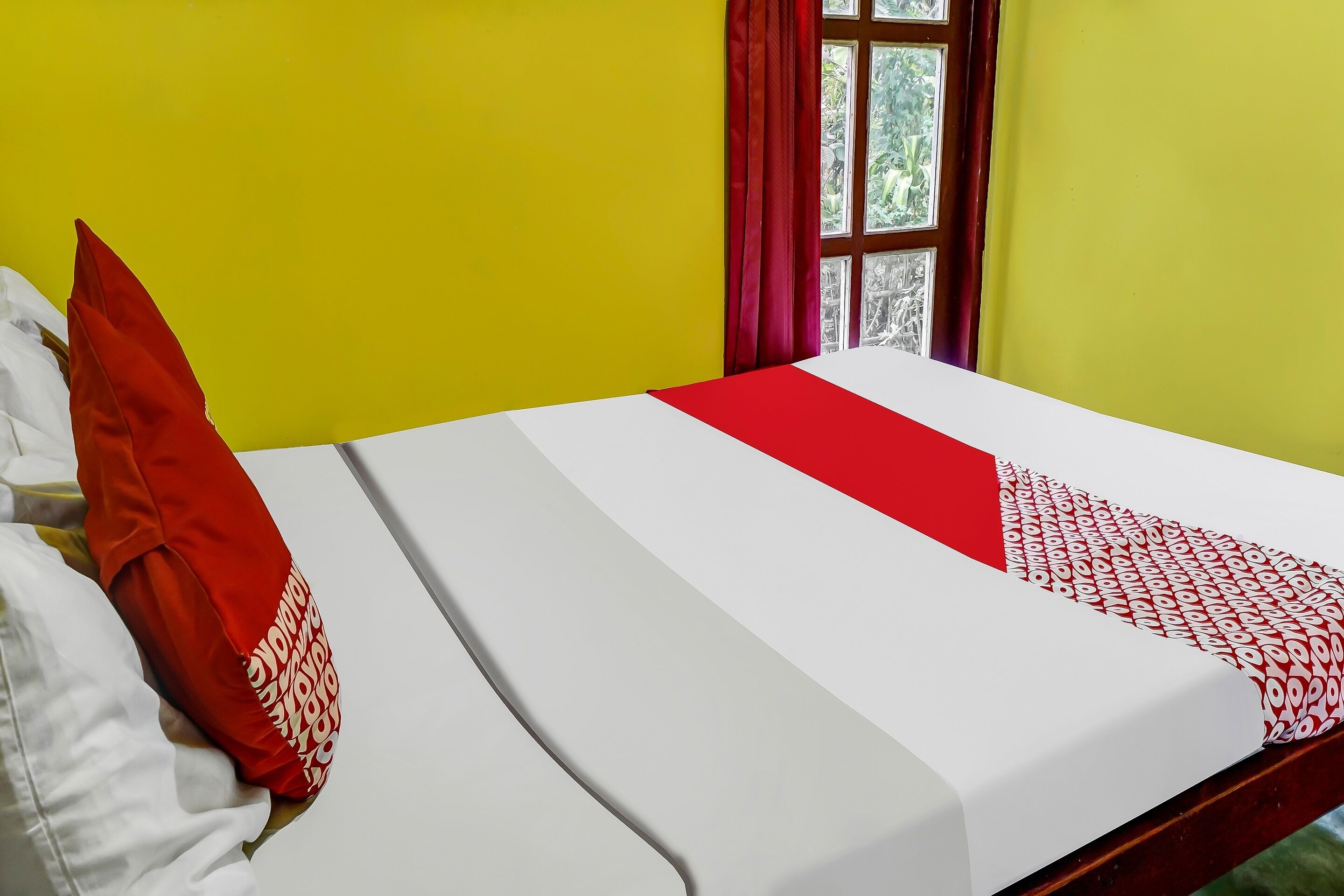 Uma cama com roupa de cama branca e vermelha, um travesseiro vermelho e uma janela com cortinas.