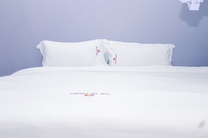 Uma cama bem arrumada com roupa de cama e almofadas brancas, uma parede lisa e um pequeno texto no cobreleito.