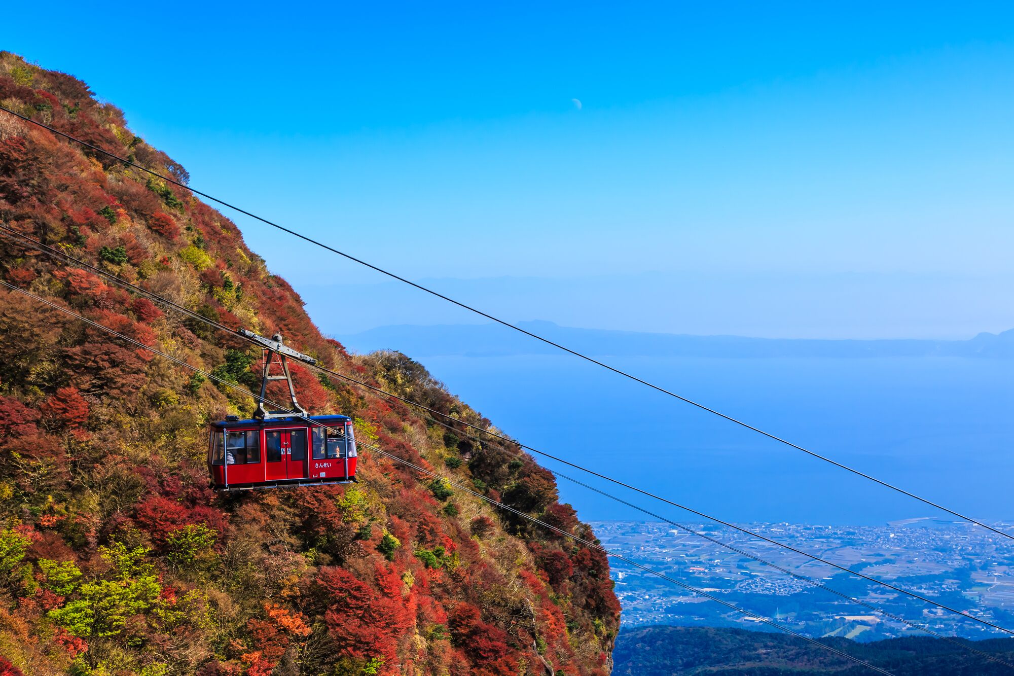 Um teleférico em movimento ao longo de uma encosta íngreme e arborizada, com vista para uma cidade distante.